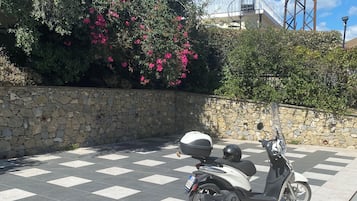 Uma scooter estacionada em um piso xadrez, com um muro de pedra e arbustos floridos ao fundo.