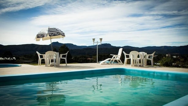 Piscina com água azul cristalina, rodeada por cadeiras de plástico brancas e um guarda-sol, com montanhas ao fundo.