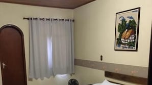 Quarto de hotel com uma cama, porta de madeira, janela com cortinas, ventoinha na parede e um quadro.