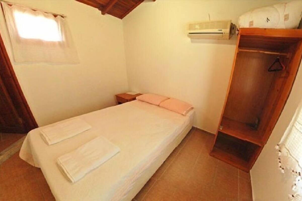 Uma cama de solteiro com roupa de cama branca, um guarda-roupa de madeira, uma pequena mesa de cabeceira e uma janela com cortina.