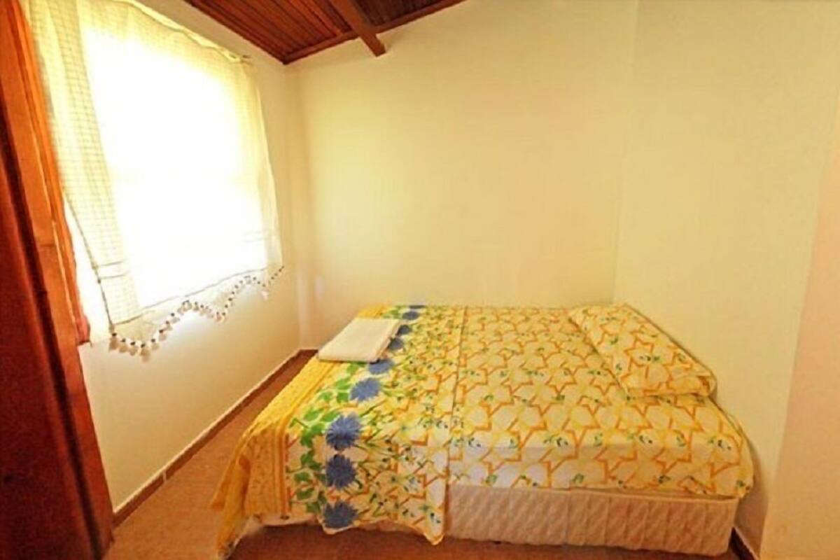 Quarto pequeno com uma cama de solteiro, parede branca simples e janela com cortina.