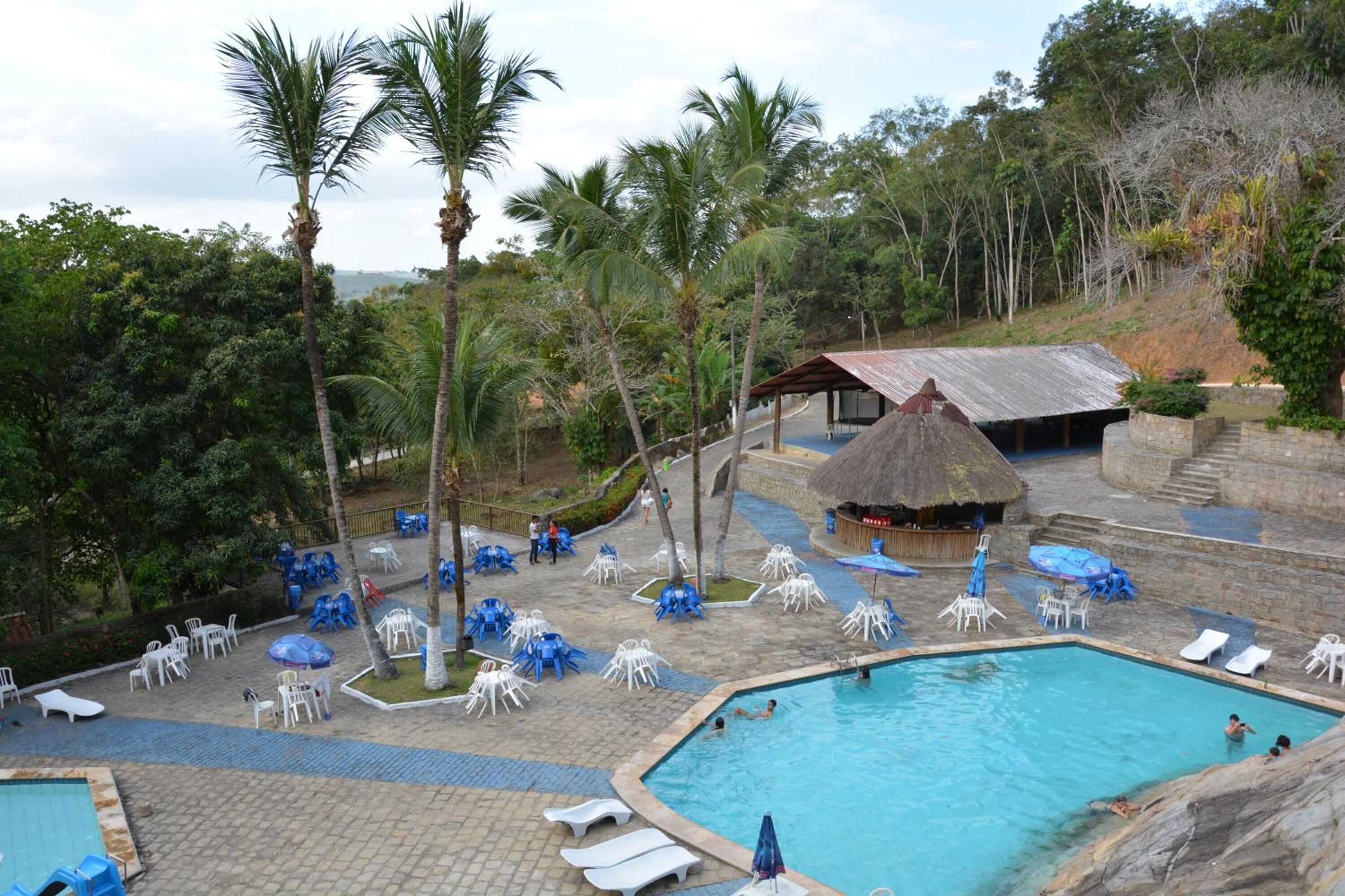 Uma área de piscina com espreguiçadeiras e guarda-sóis, cercada por palmeiras e uma construção com telhado de palha.