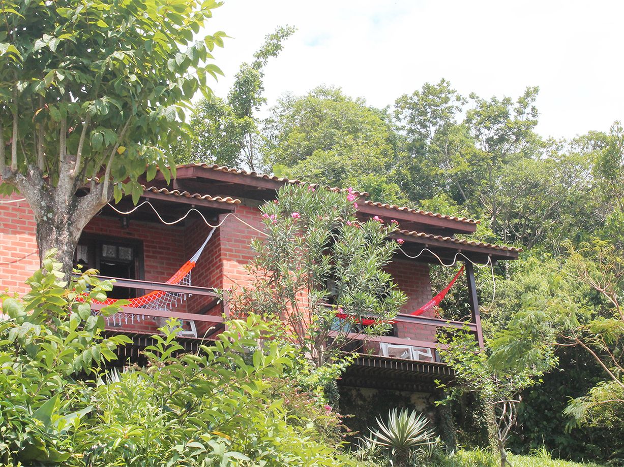 Um edifício de dois andares em tijolo com varanda, rodeado por vegetação exuberante.