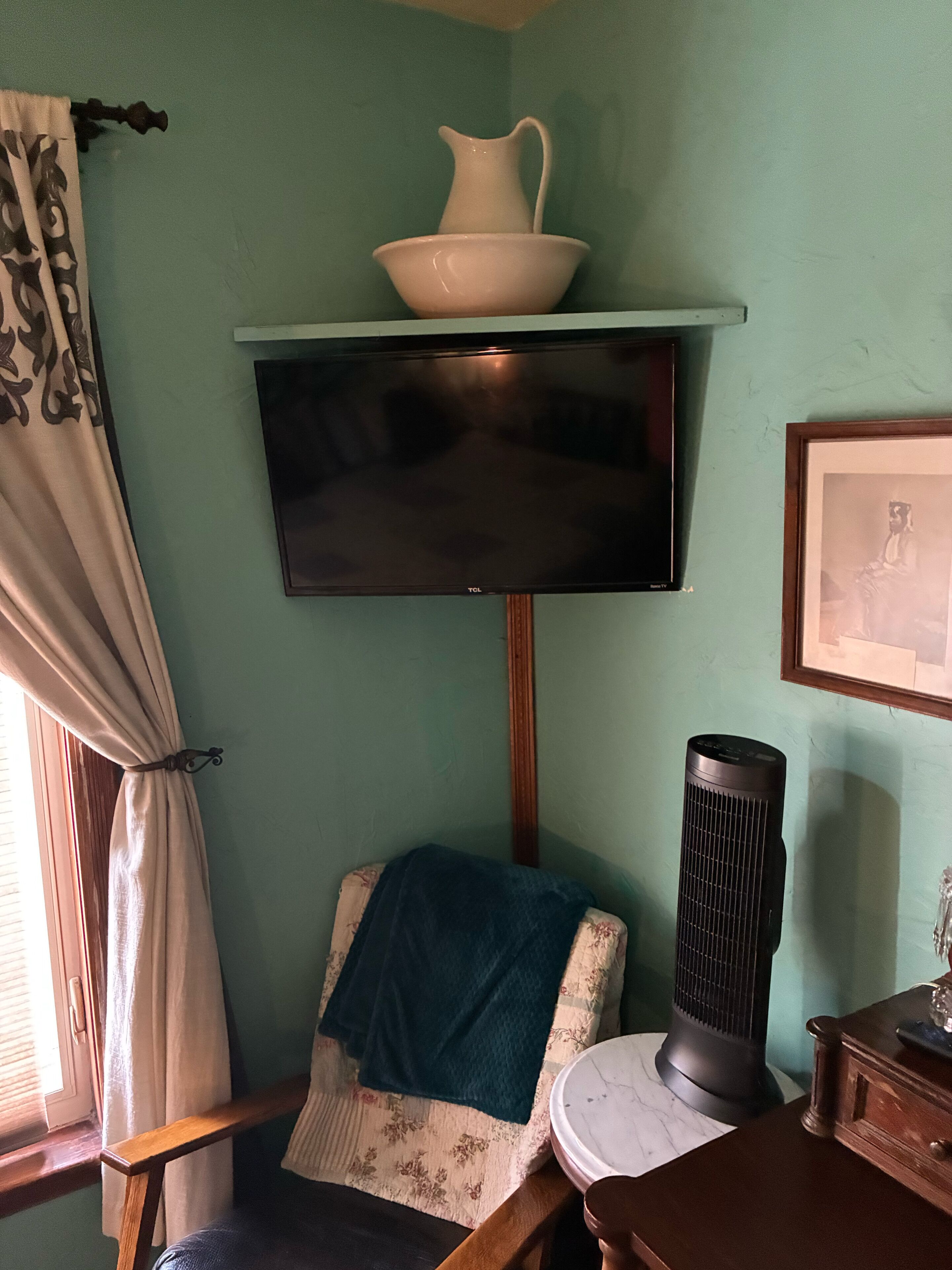 Quarto com televisão na parede, uma cadeira com manta, radiador e janela com cortinas.
