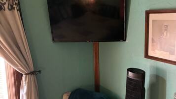 Quarto com televisão na parede, uma cadeira com manta, radiador e janela com cortinas.