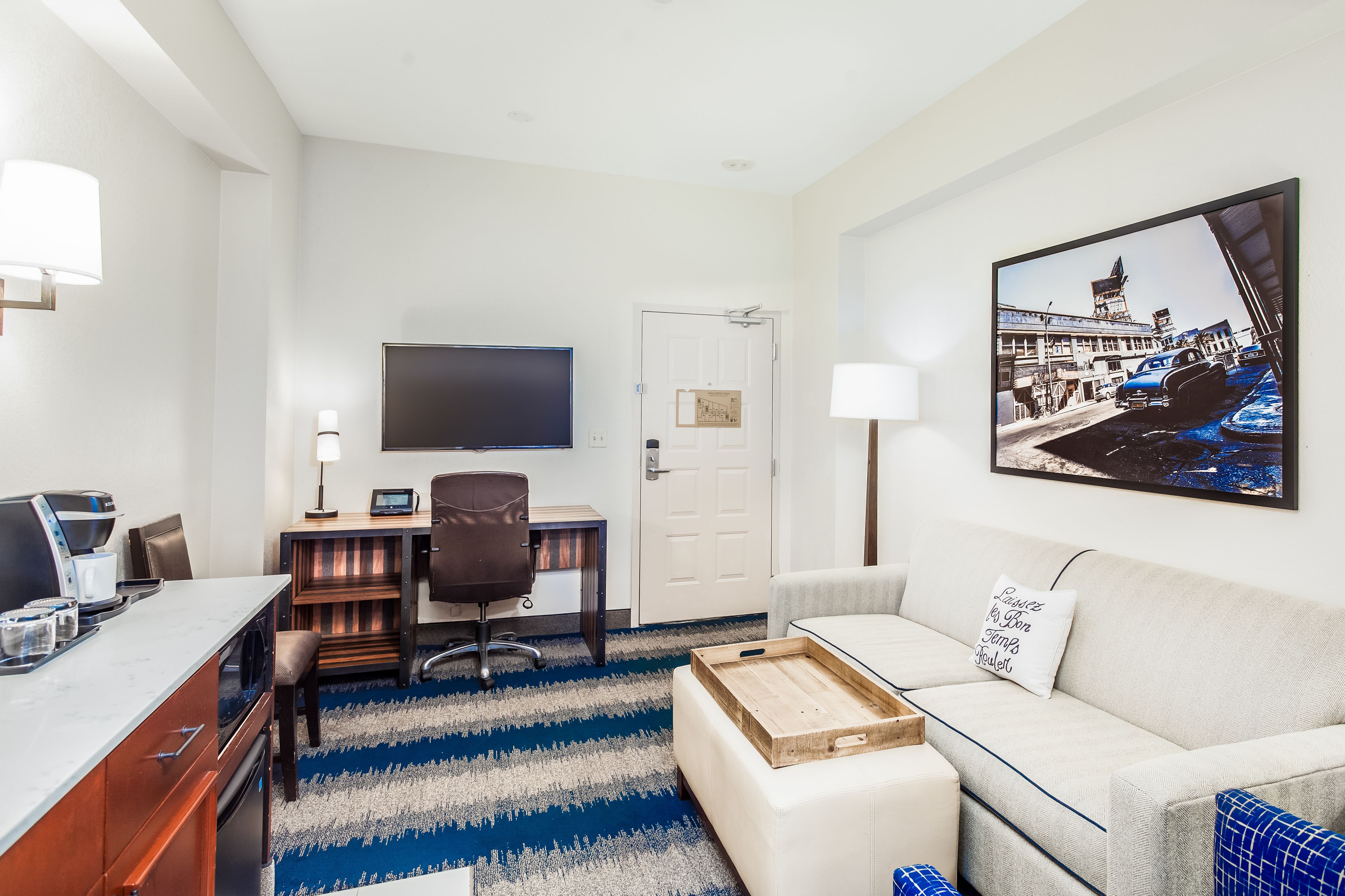 Quarto de hotel com televisão de tela plana, uma escrivaninha com computador, uma cadeira, um sofá, uma mesa de centro e uma grande moldura na parede.
