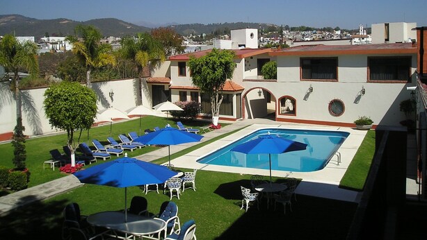 Uma área de piscina com espreguiçadeiras e guarda-sóis, rodeada por um gramado bem cuidado e um edifício com telhado de telhas vermelhas.