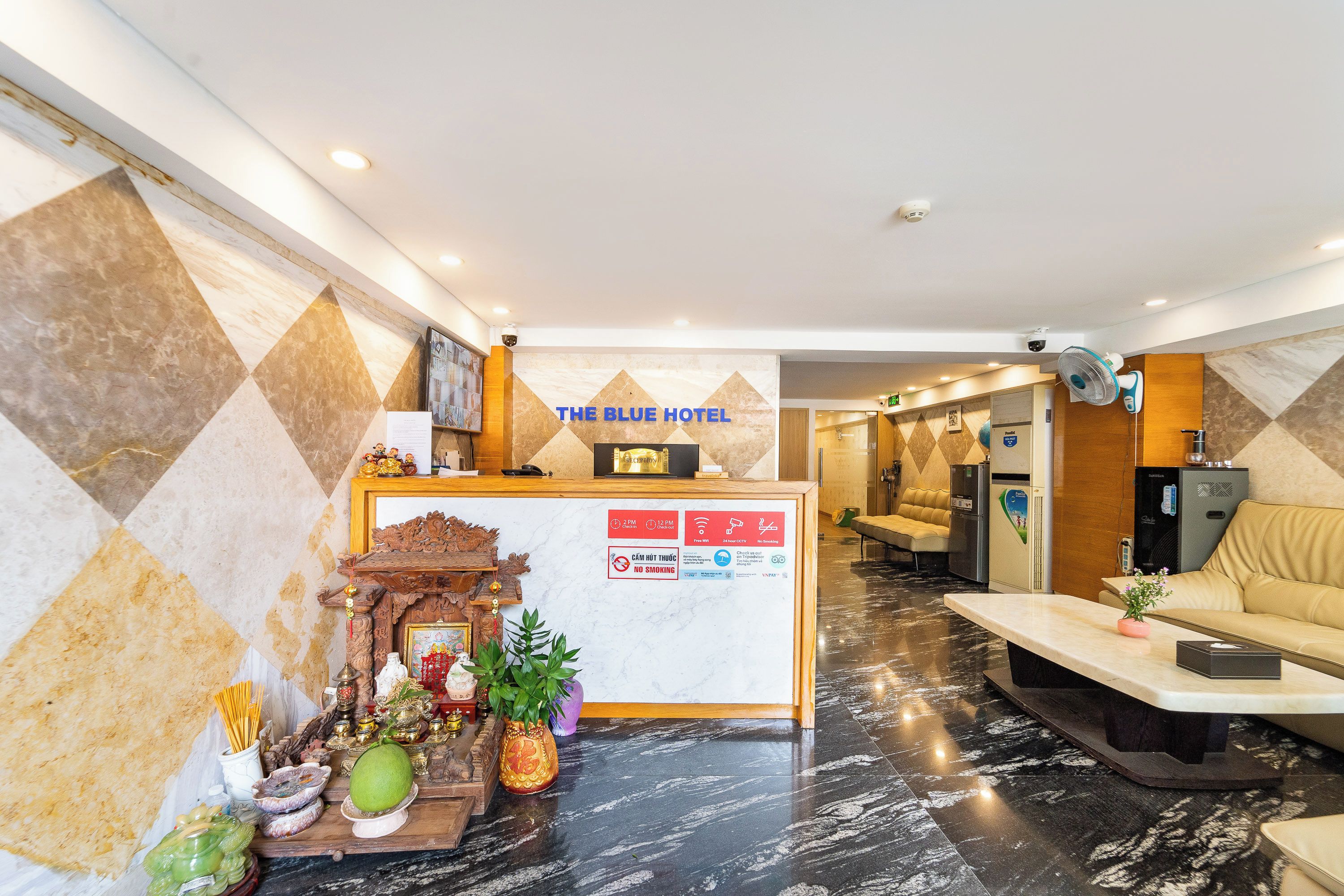 Área de recepção de hotel com balcão, câmera de segurança na parede, sofá, geladeira e um altar decorativo com oferendas.