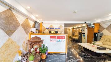 Área de recepção de hotel com balcão, câmera de segurança na parede, sofá, geladeira e um altar decorativo com oferendas.