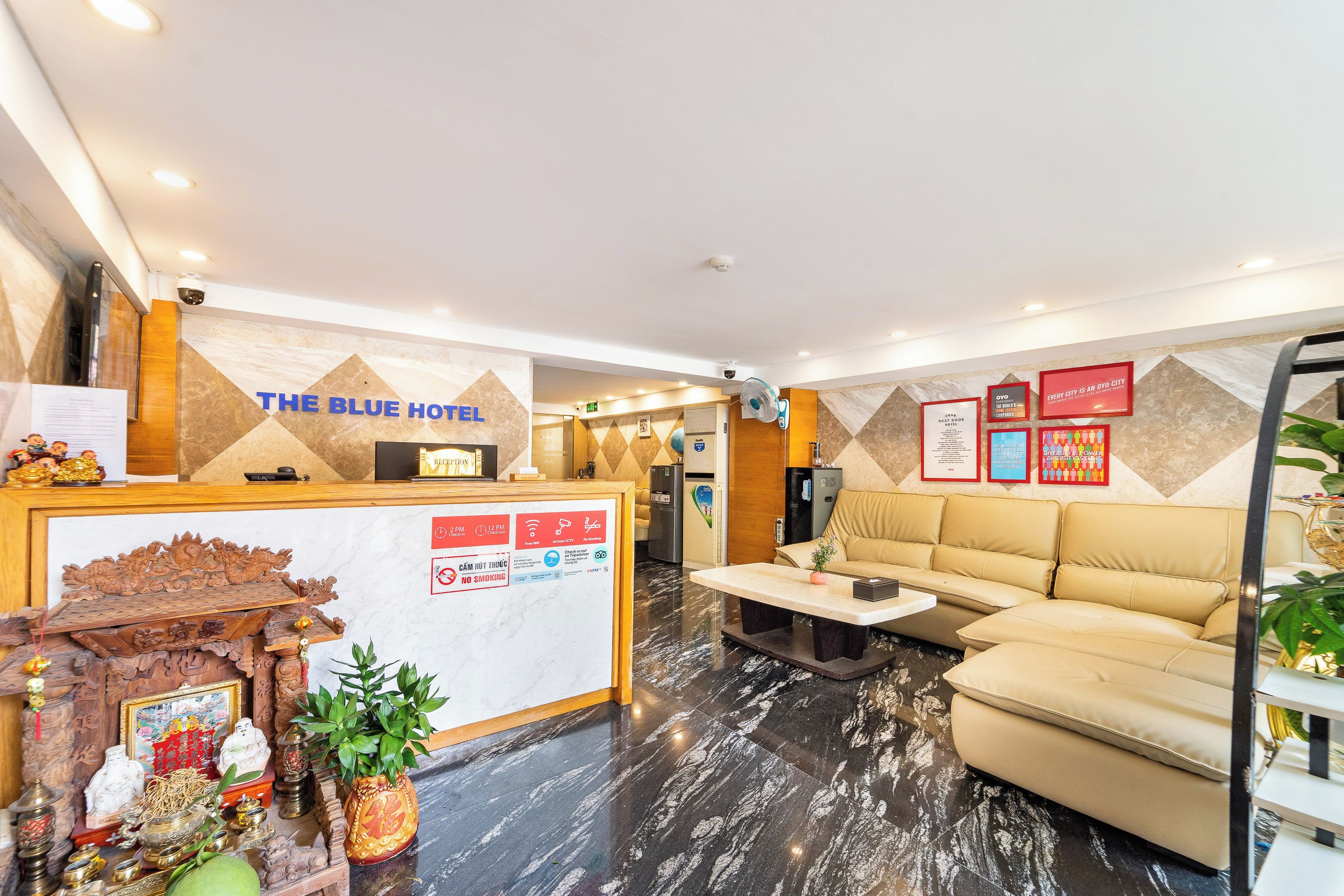 Uma área de recepção de hotel com balcão, sofá e mesa de centro. Há placas indicando horários de check-in e política de não fumantes.