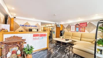 Uma área de recepção de hotel com balcão, sofá e mesa de centro. Há placas indicando horários de check-in e política de não fumantes.