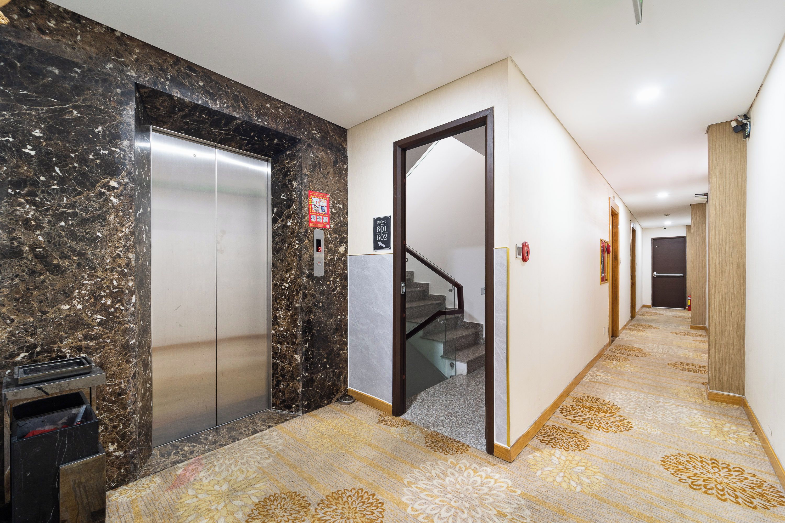 Corredor de hotel com parede de mármore, um elevador, uma escadaria e piso coberto de carpete.