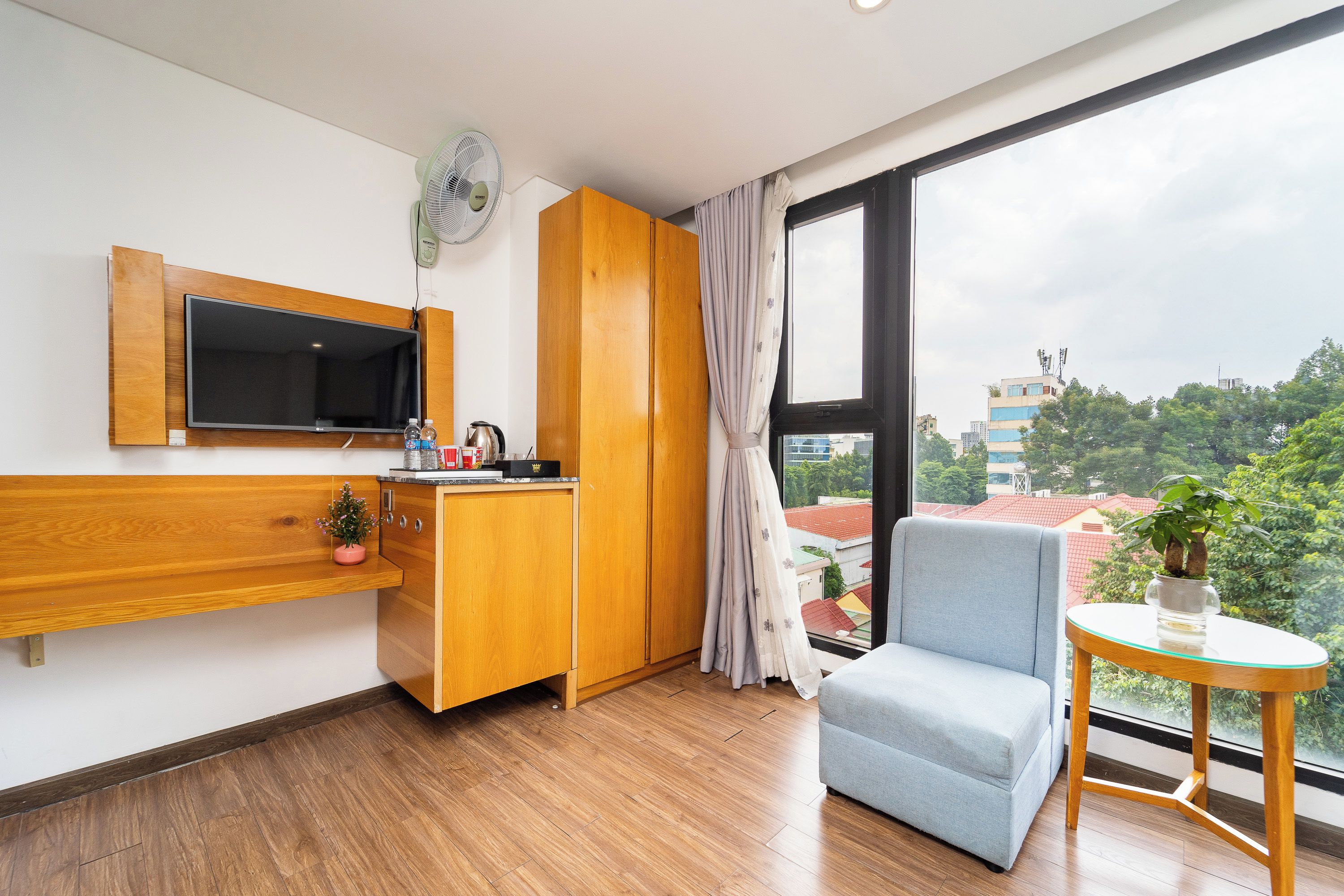 Quarto moderno com armário de madeira, TV de tela plana, pequena mesa com planta em vaso, sofá cinza e grande janela com vista para a cidade.