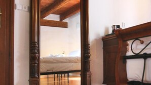 Quarto com piso de madeira, um espelho grande, uma cama com roupa de cama branca e uma mesa pequena com estrutura de metal.