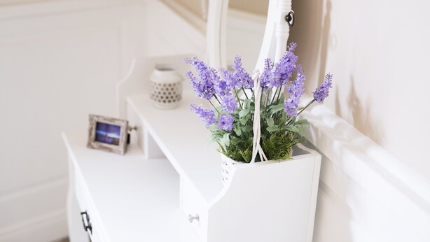 Uma toucador branca com espelho, uma fotografia pequena emoldurada e um vaso com flores roxas.