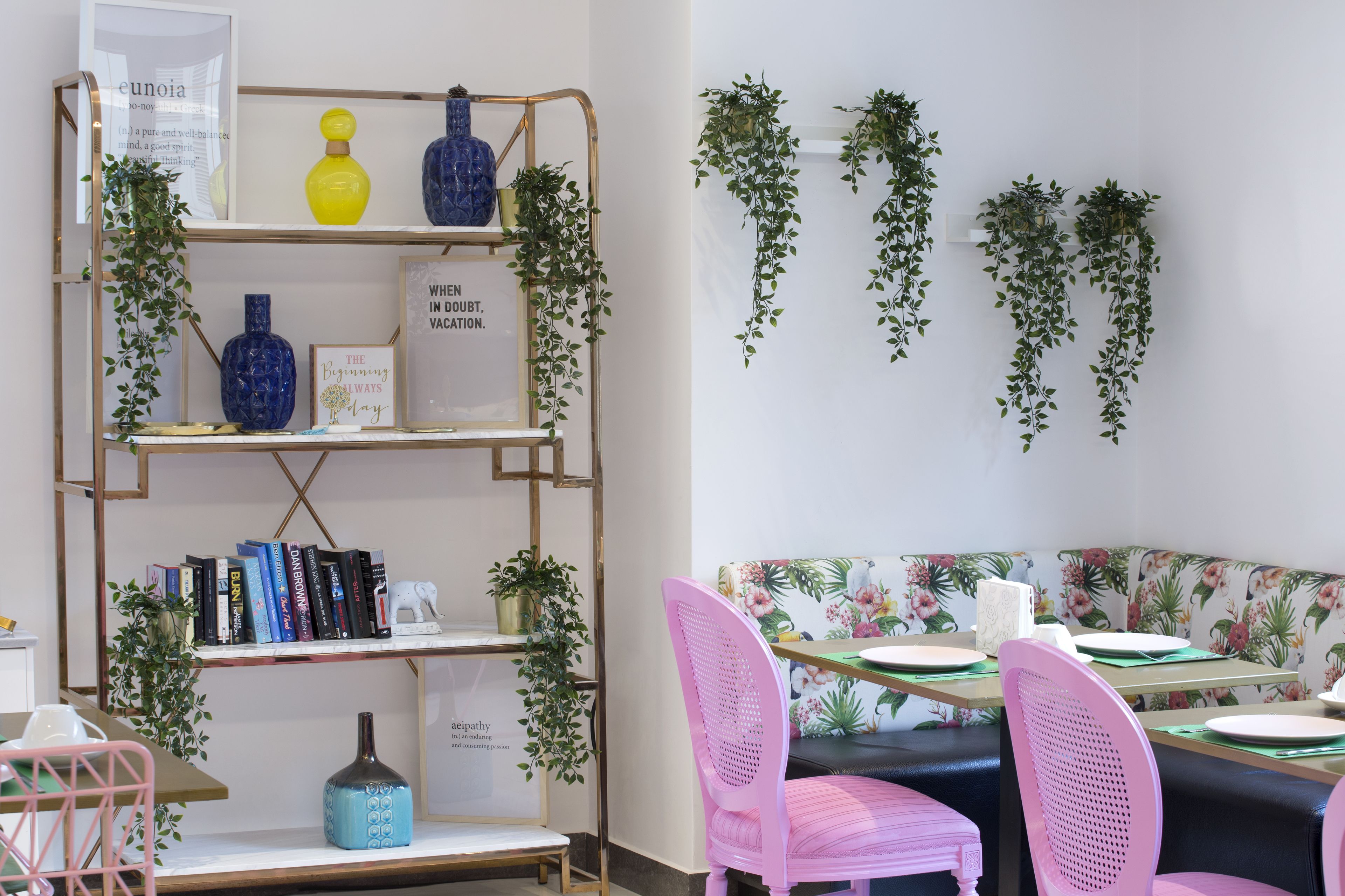 Um espaço de estar acolhedor com um banco estampado em floral e cadeiras cor-de-rosa, uma estante com vasos decorativos e livros, e plantas pendentes na parede.