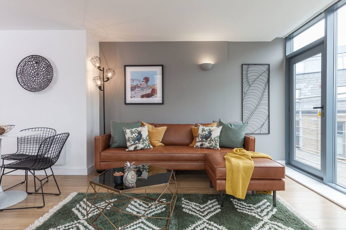 Uma sala de estar moderna com um sofá de couro marrom, almofadas verdes e amarelas, uma mesa de centro de vidro e uma janela grande com vista para a cidade.