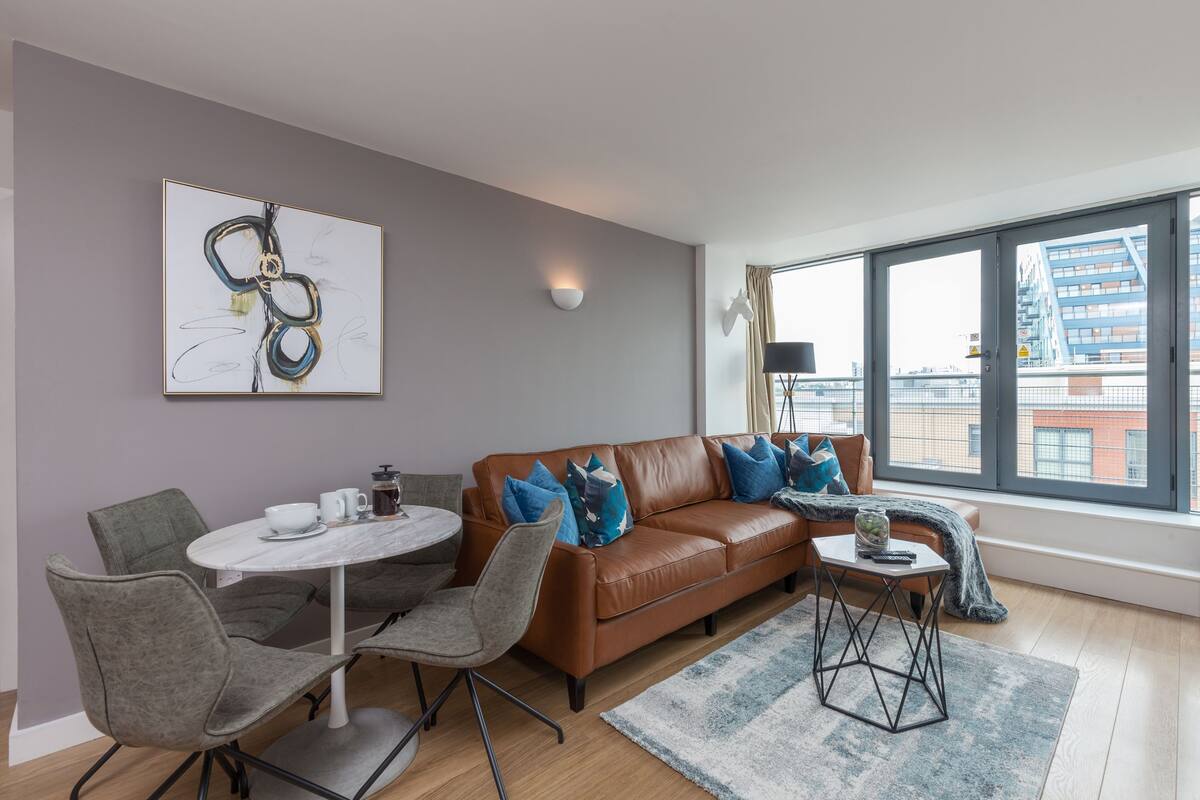 Uma sala de estar moderna com um sofá de couro marrom, poltronas cinzentas, uma mesa de centro redonda e uma grande janela com vista para edifícios.