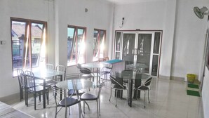 Uma área de refeições com mesas redondas de vidro e cadeiras de metal, janelas amplas com cortinas e vista para o exterior.