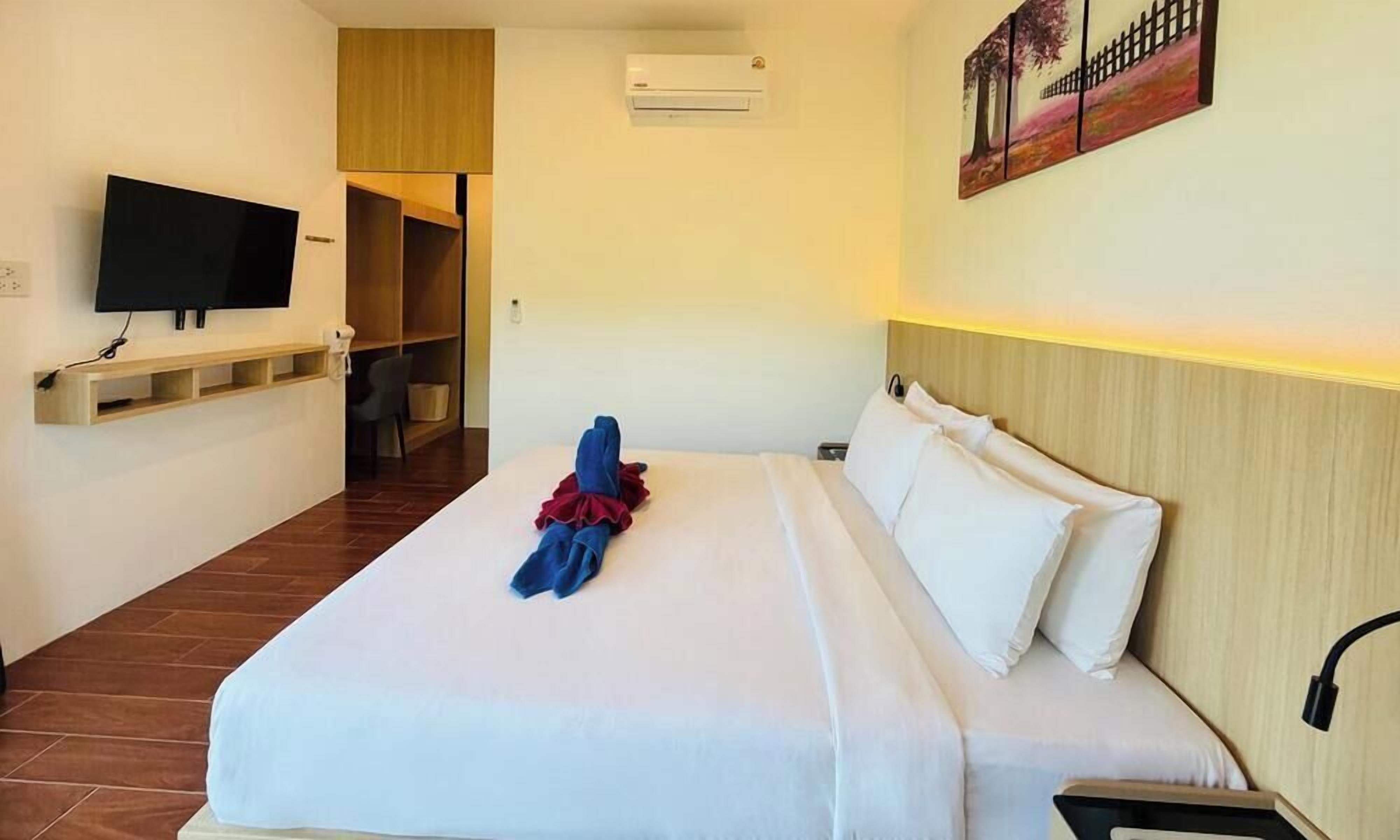 Quarto de hotel com cama, televisão de tela plana na parede, um quadro e cabeceira de madeira.