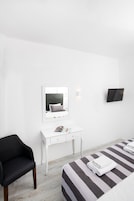Quarto de hotel minimalista com uma cama, uma escrivaninha branca, uma cadeira preta, televisão de tela plana e um espelho.