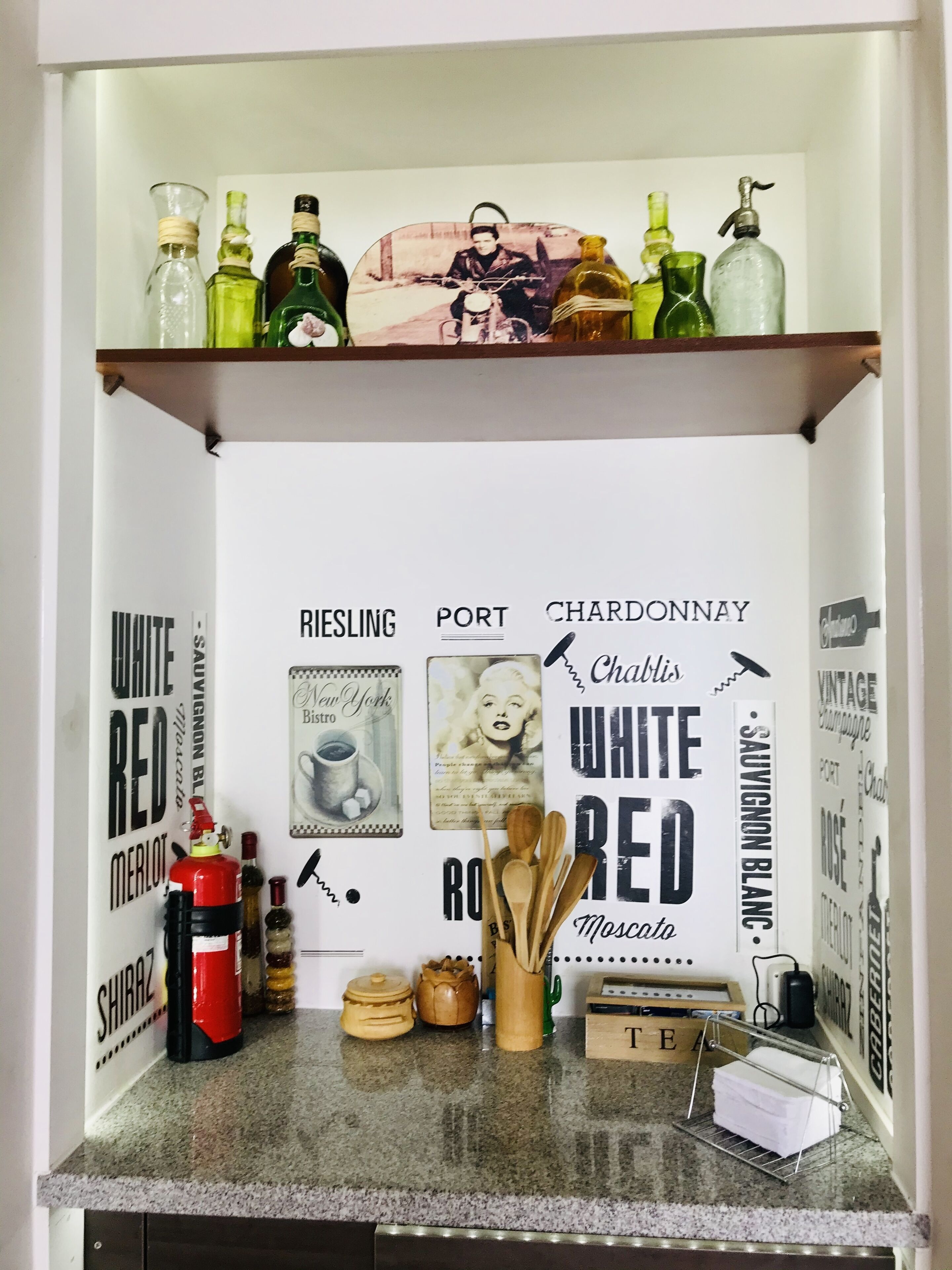 Uma bancada de cozinha com vários frascos, um extintor de incêndio vermelho, utensílios de madeira e uma lousa com texto sobre vinho.