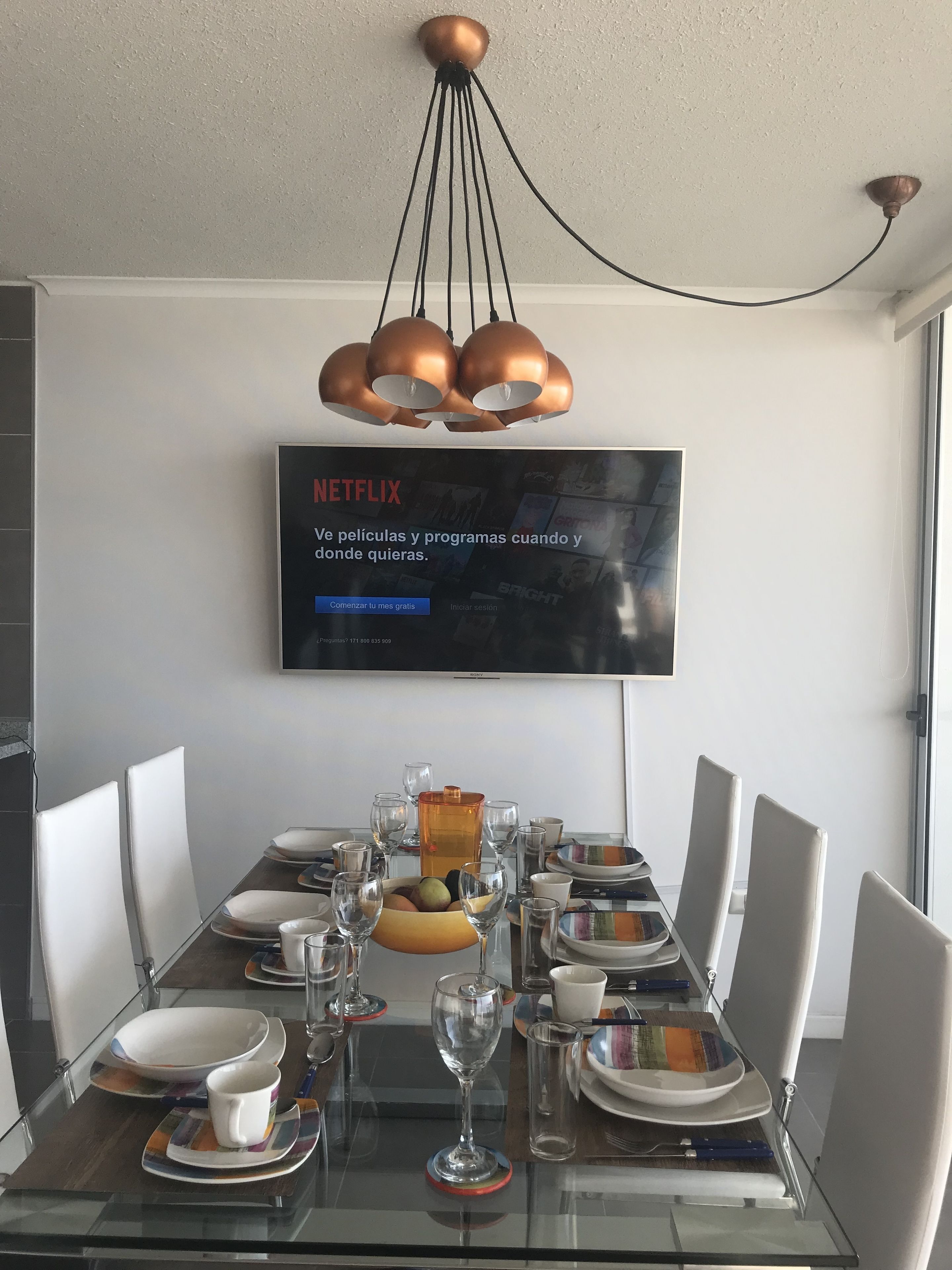 Uma sala de jantar moderna com uma mesa de vidro posta para seis pessoas, cadeiras brancas e uma televisão na parede exibindo a interface da Netflix.