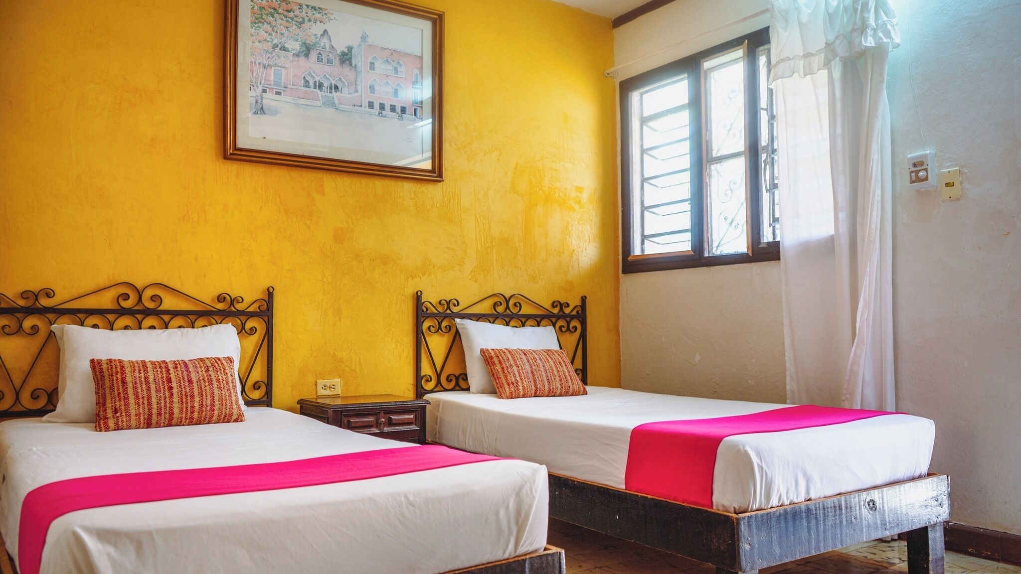 Duas camas com estrutura de metal, roupa de cama branca e detalhes vermelhos, num quarto com paredes amarelas e uma janela com cortinas brancas.