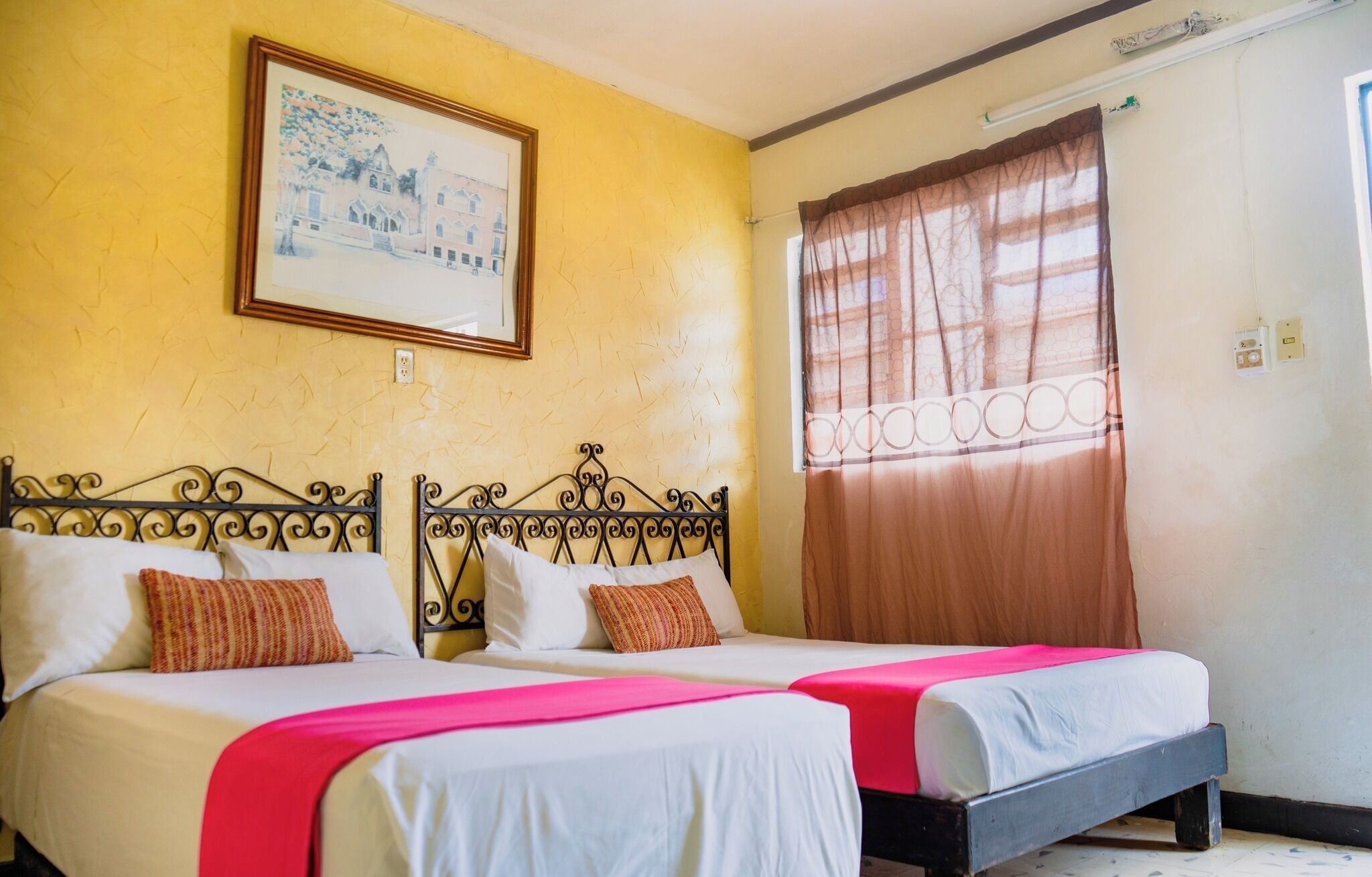 Um quarto com duas camas, um quadro pendurado na parede, uma janela com cortinas e um termostato na parede.
