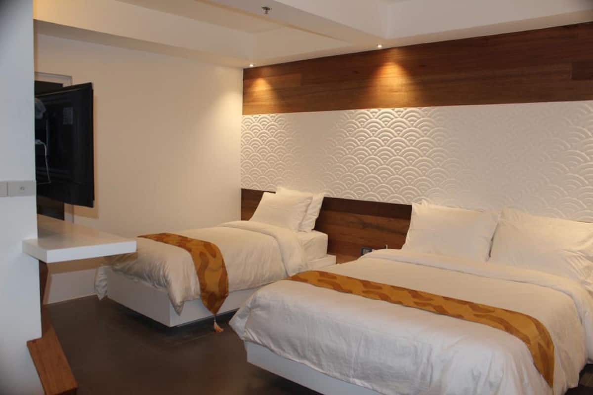 Quarto de hotel moderno com duas camas, painel de cabeceira de madeira e TV integrada.