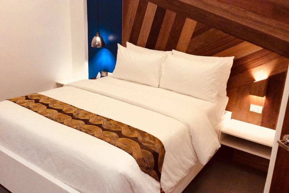 Cama bem arrumada com lençóis brancos e manta estampada, mesa de cabeceira com abajur, e estrutura de teto em madeira.