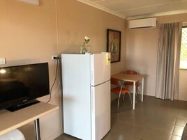 Quarto compacto com TV de tela plana, geladeira branca, uma pequena mesa com duas cadeiras e aparelho de ar condicionado.