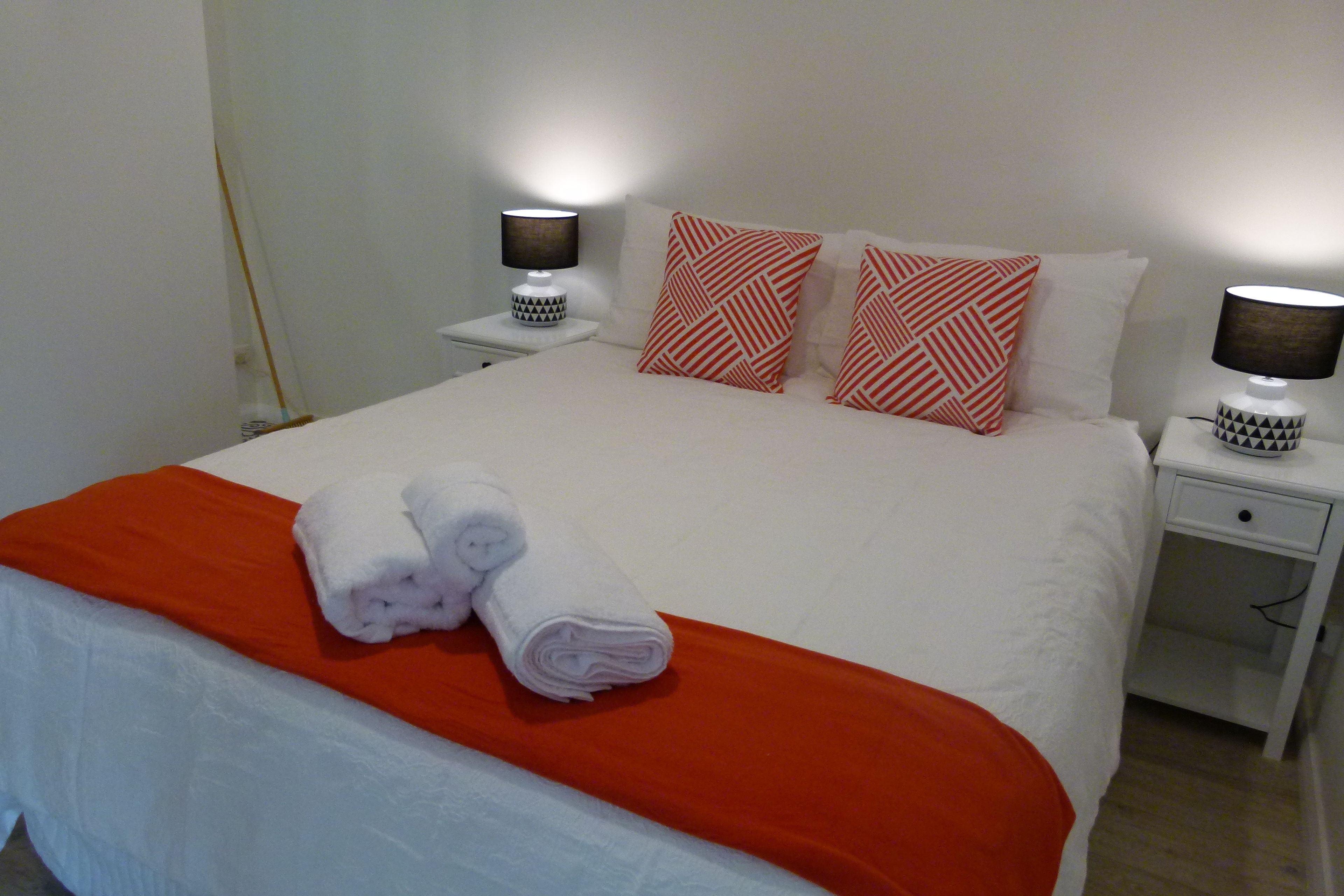 Cama bem arrumada com roupa de cama branca e laranja, dois travesseiros estampados e duas luminárias de cabeceira em mesas de cabeceira brancas.