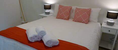 Cama bem arrumada com roupa de cama branca e laranja, dois travesseiros estampados e duas luminárias de cabeceira em mesas de cabeceira brancas.