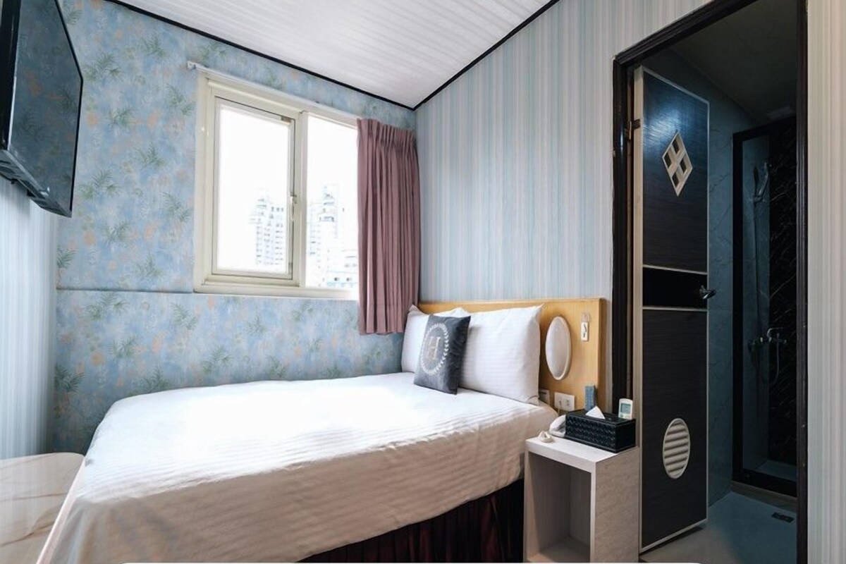 Quarto de hotel com cama, mesa de cabeceira, janela com cortinas e porta com puxador.