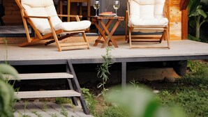Uma cabana de madeira com varanda, duas cadeiras estofadas, uma mesinha com um copo de vinho e uma escada encostada na parede.