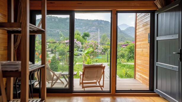 Quarto com decoração em madeira, porta de vidro deslizante para uma varanda com cadeira e mesa, e vista para uma paisagem montanhosa.