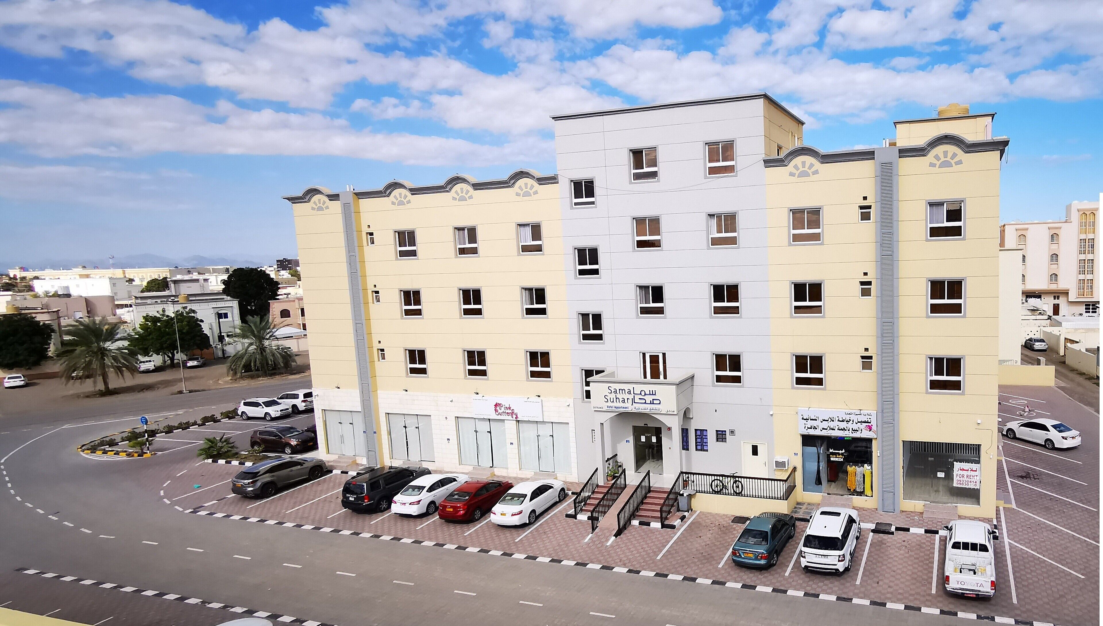 Um hotel de vários andares com entrada central, estacionamento e edifícios ao redor.