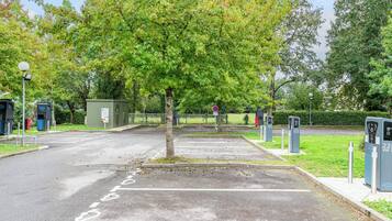 Área de estacionamento com estações de carregamento para veículos elétricos, uma árvore e uma cerca.
