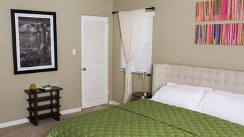 Quarto com colcha verde acolchoada, fotografia a preto e branco emoldurada, porta branca, mesa de cabeceira com abajur e obra de arte colorida com padrão listrado na parede.