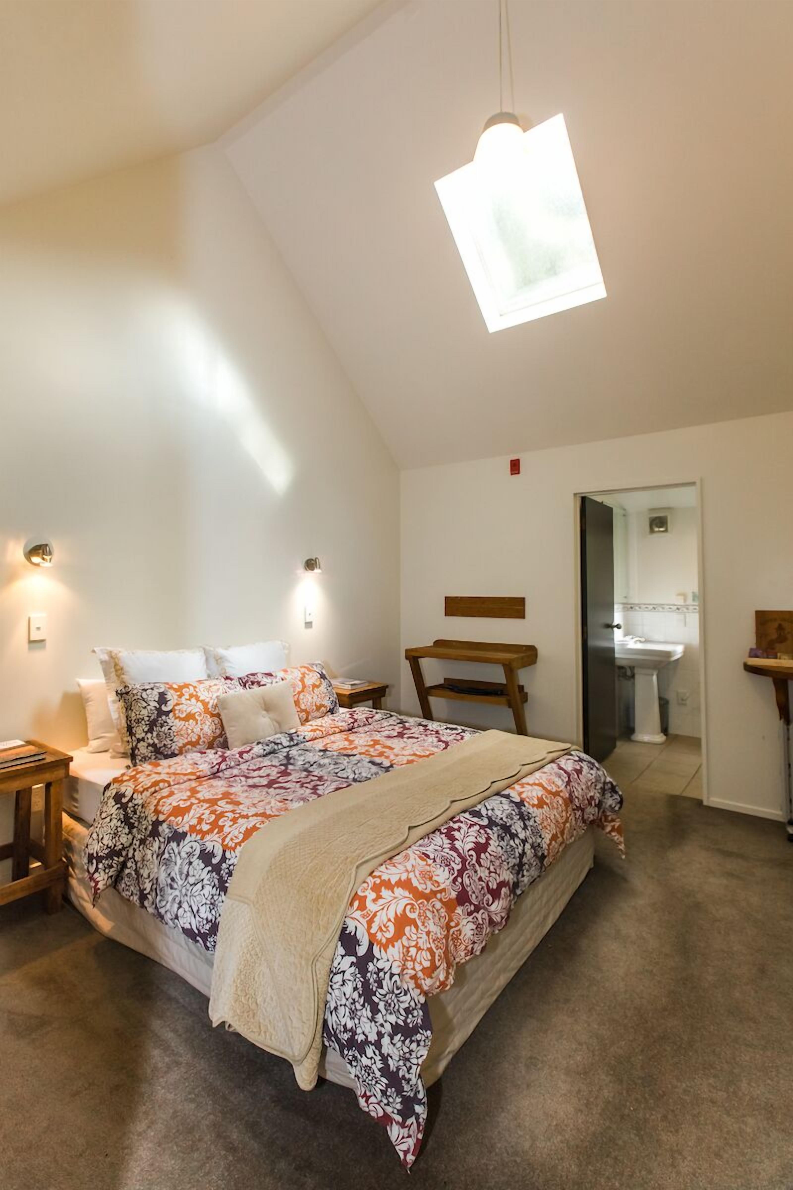 Quarto com uma cama grande, mesas de cabeceira, um banquinho, um candeeiro pendente e um banheiro visível através de uma porta aberta.