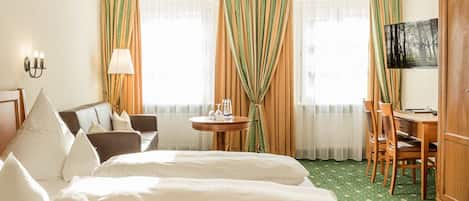 Quarto de hotel com uma cama grande, uma secretária com cadeiras, uma televisão e um candelabro.