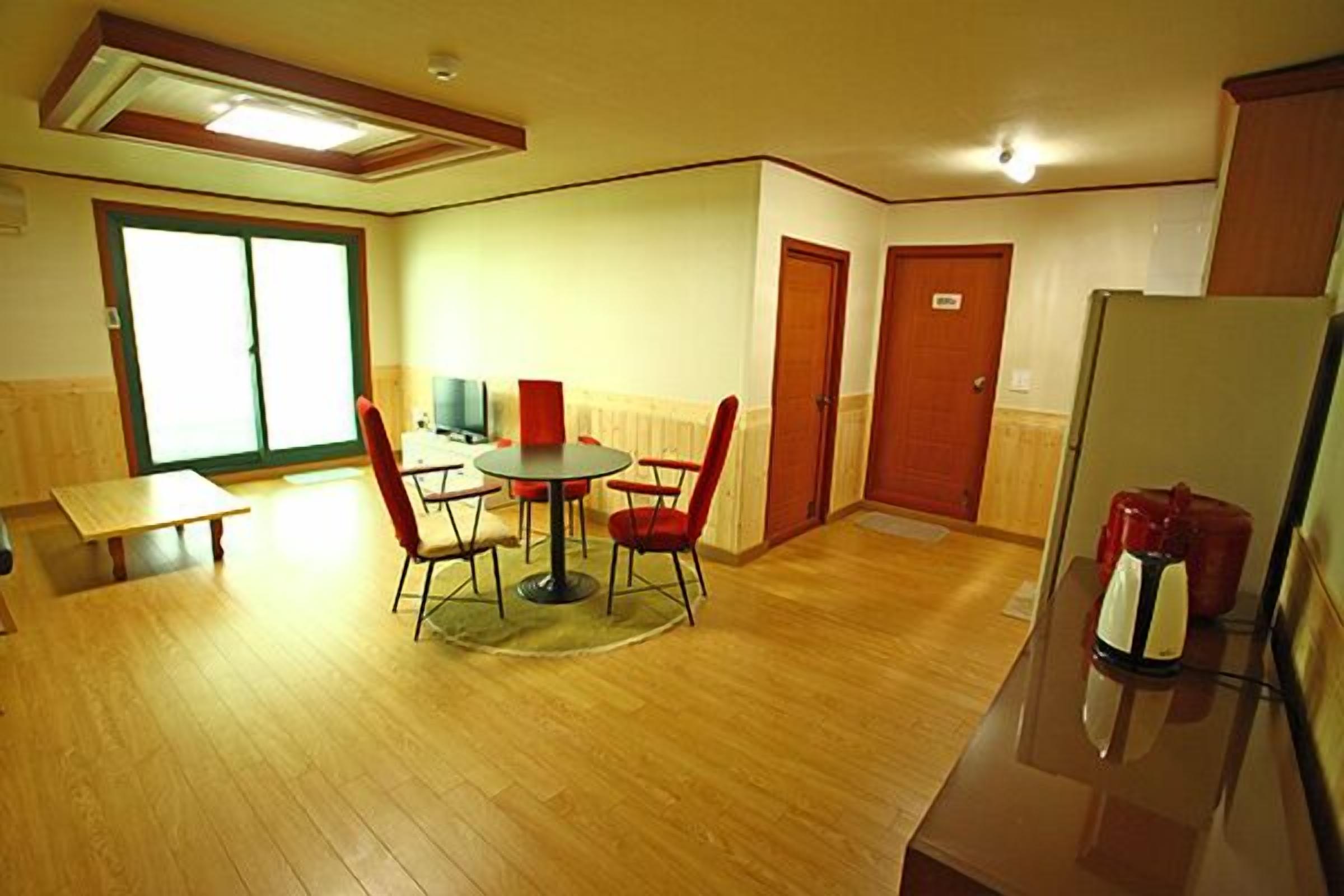 Um quarto com piso de madeira, uma mesa redonda com quatro cadeiras, um banco, uma geladeira e uma porta deslizante.