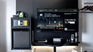 Quarto de hotel com uma grande tela de TV exibindo o nome do hotel e uma vista urbana. Há uma pequena geladeira, uma máquina de café e uma prateleira com diversos objetos. Duas cadeiras brancas estão posicionadas em frente a uma mesa.