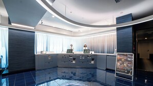 Um lobby de hotel moderno com um balcão de recepção curvo, um painel de informações digital e uma grande janela com persianas.
