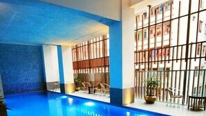 Piscina interna com água azul cristalina, rodeada por paredes com ripas verticais de madeira e vista para edifícios externos.
