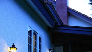 Uma casa com fachada branca, janela com moldura branca e chaminé de tijolos. Uma escadaria de madeira leva a um alpendre coberto.