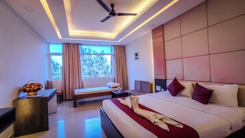 Quarto de hotel moderno com uma cama grande, uma escrivaninha e uma cadeira. Há uma janela com cortinas, um ventilador de teto e vista para árvores.