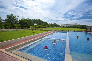Área de piscina com várias piscinas, gramado com árvores e um edifício ao fundo.