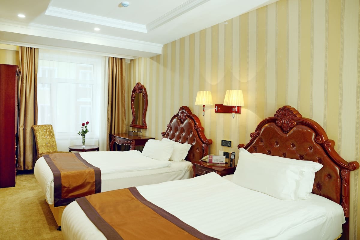 Quarto de hotel com duas camas, um espelho grande, mesa de cabeceira com abajur e janela com cortinas.